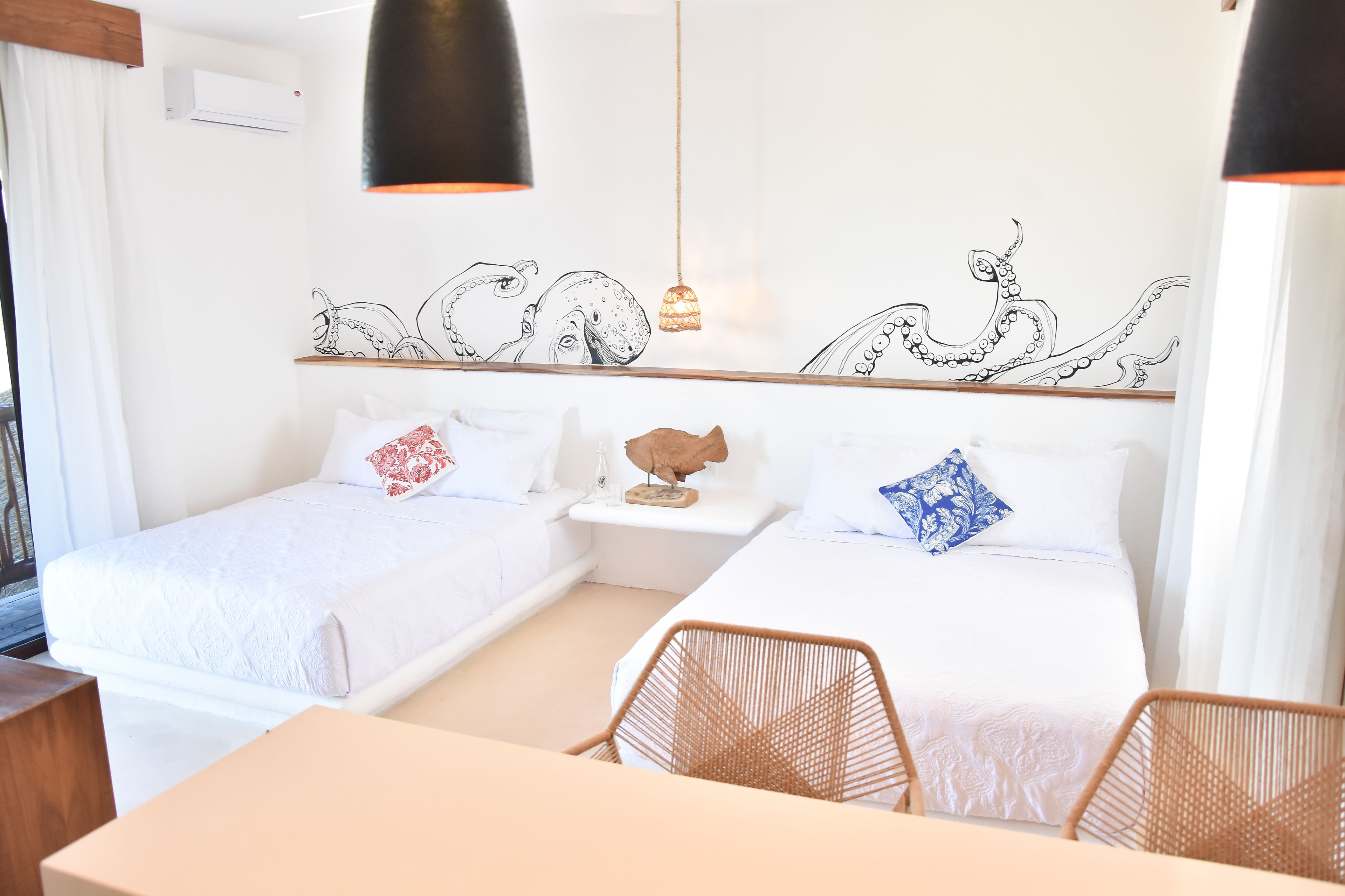Quarto com duas camas, uma mesa pequena, cadeiras de vime e uma parede com ilustrações de polvos.