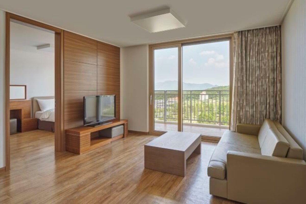 Uma sala de estar moderna com piso de madeira, uma TV de tela plana sobre um suporte de madeira, um sofá bege e um grande vão de vidro deslizante que dá para uma varanda com vista para áreas verdes e montanhas.