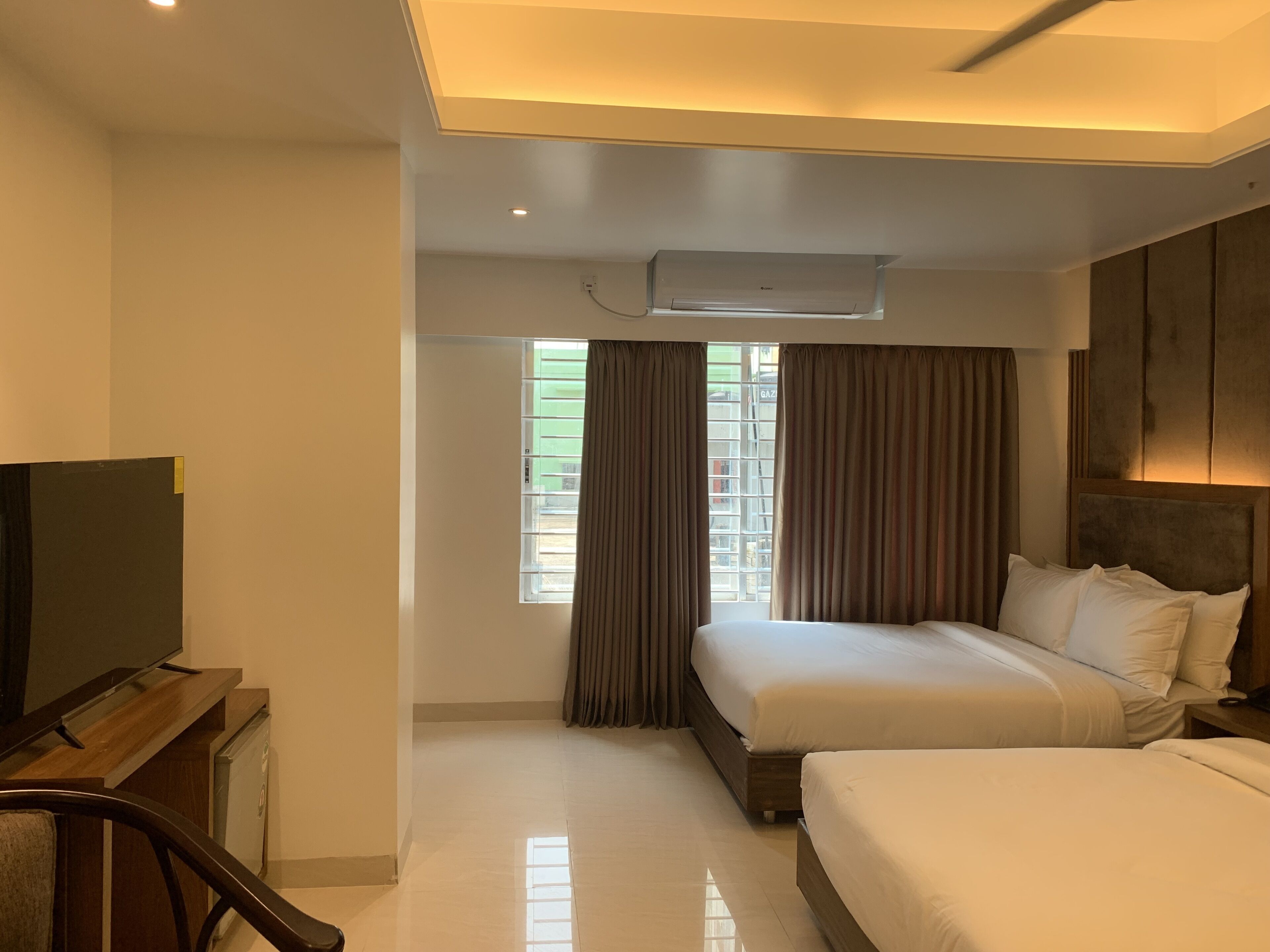 Quarto de hotel com duas camas, televisão, ventoinha no teto e janela com cortinas.