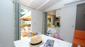 Quarto com uma mesa branca, um chapéu e uma revista. Há uma janela com cortinas, uma parede branca e uma porta que dá para outro quarto.