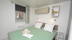 Quarto arrumado com uma cama de casal, janela com cortinas, prateleira na parede e uma mesinha de cabeceira.