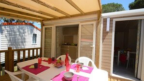 Uma área de refeições coberta ao ar livre com uma mesa posta para dois, rodeada por cadeiras brancas, e uma porta de vidro deslizante que dá para uma cozinha.