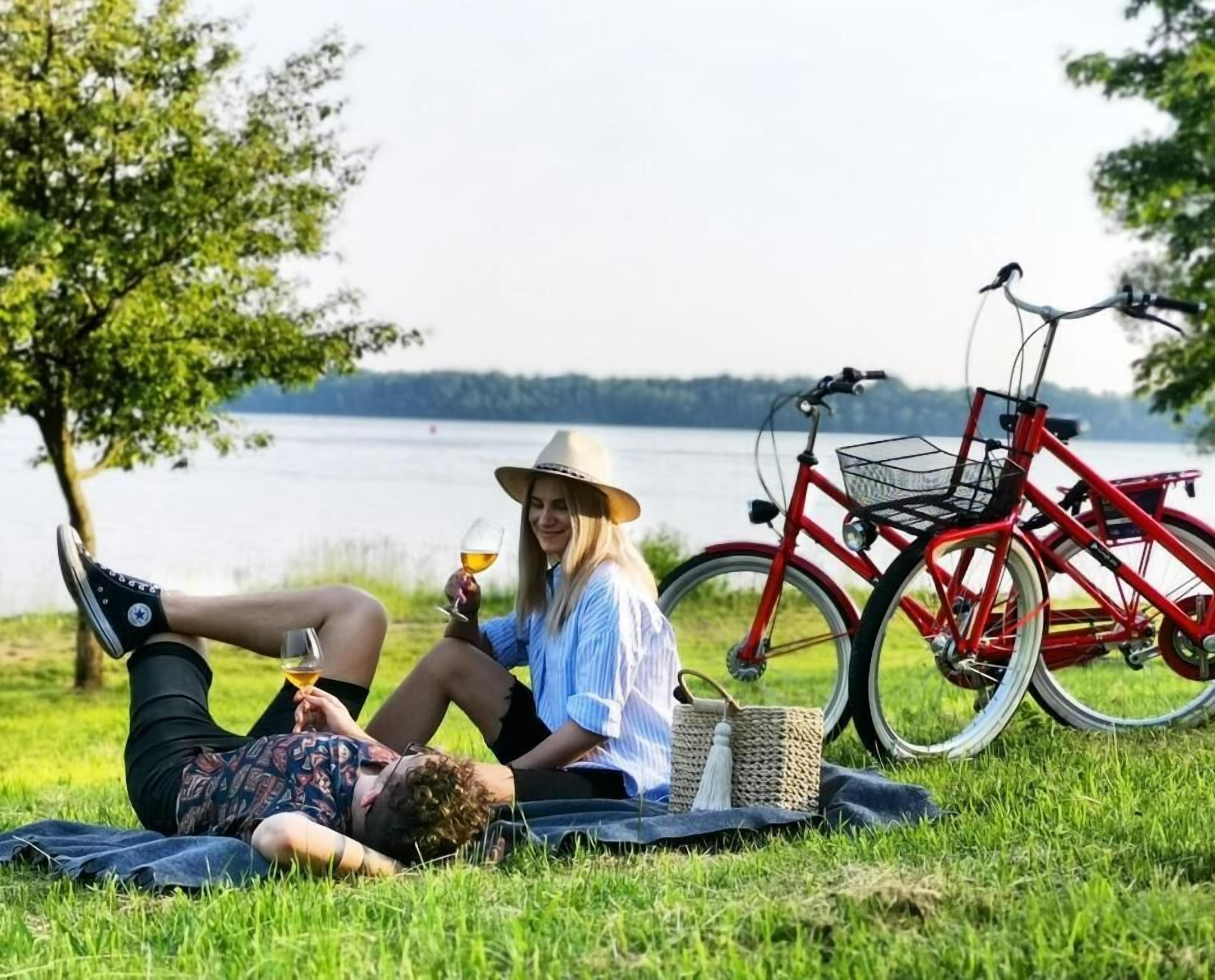 Duas pessoas relaxando em um cobertor perto de um lago, com uma bicicleta e uma cesta por perto.