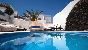 Piscina azul cristalina com cadeiras de descanso e um bangalô, cercada por edifícios brancos e palmeiras.