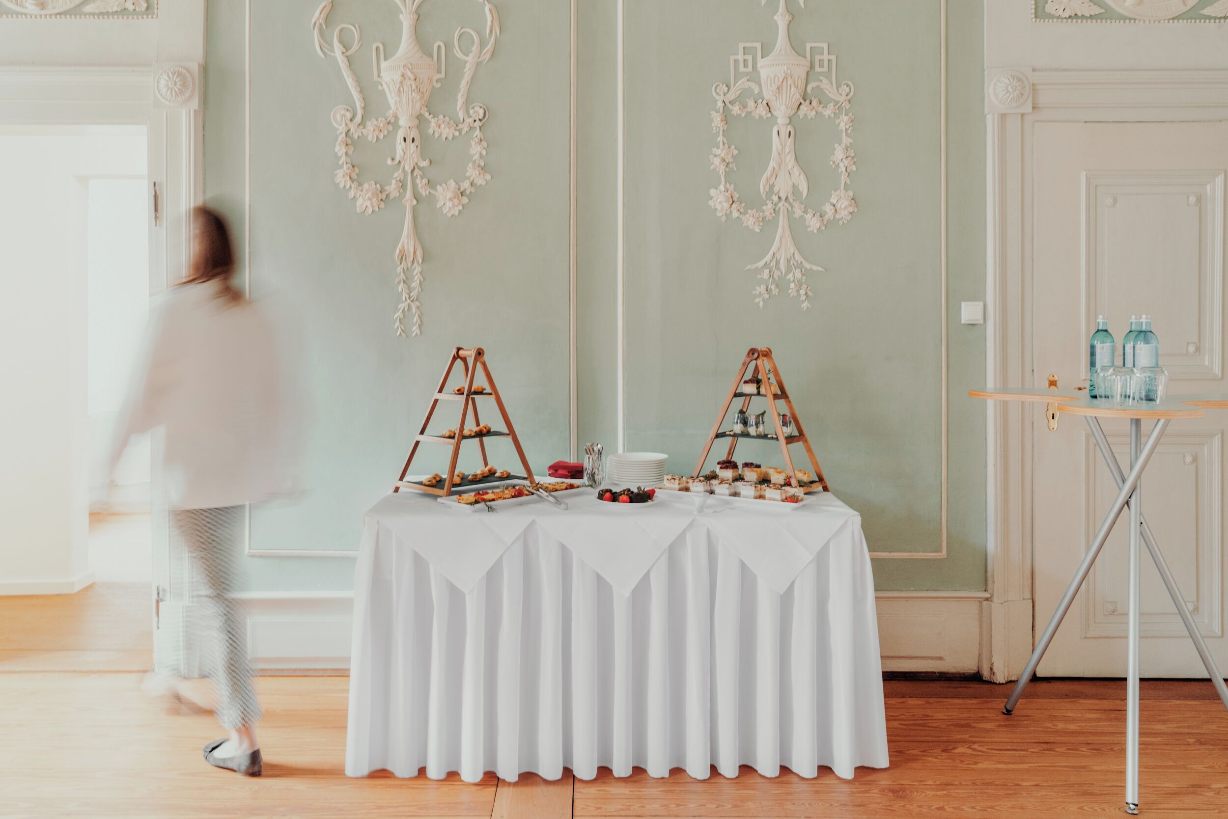 Quarto com uma mesa branca coberta por um tecido branco, dois suportes de madeira com produtos alimentícios, e uma pequena mesa com garrafas.