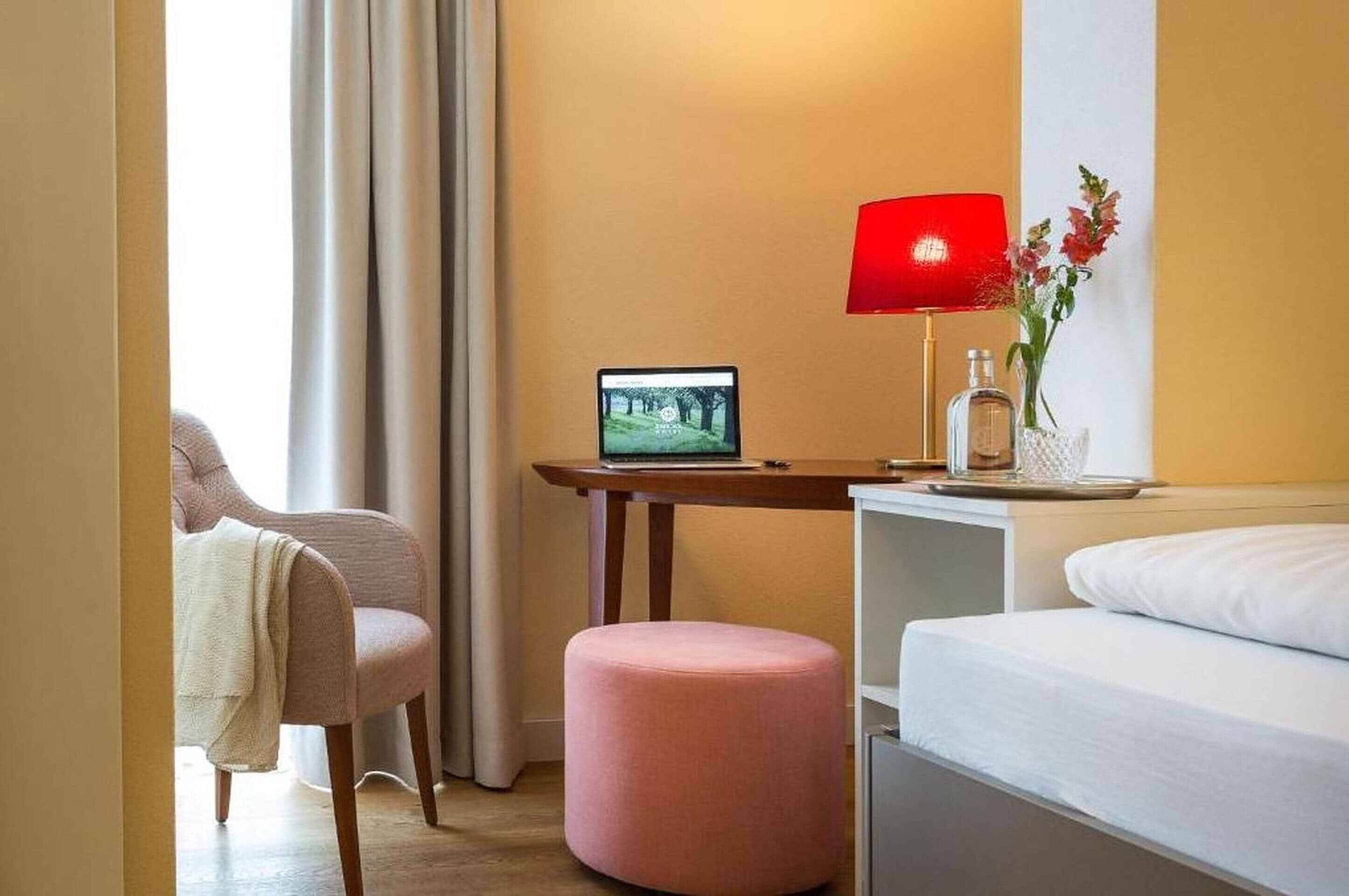 Quarto de hotel com cama, secretária com computador, cadeira e um banquinho rosa.