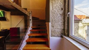 Uma escadaria com degraus de madeira e corrimão de metal, conduzindo a um quarto com parede de pedra e janela com vista para um edifício com telhado de telhas.