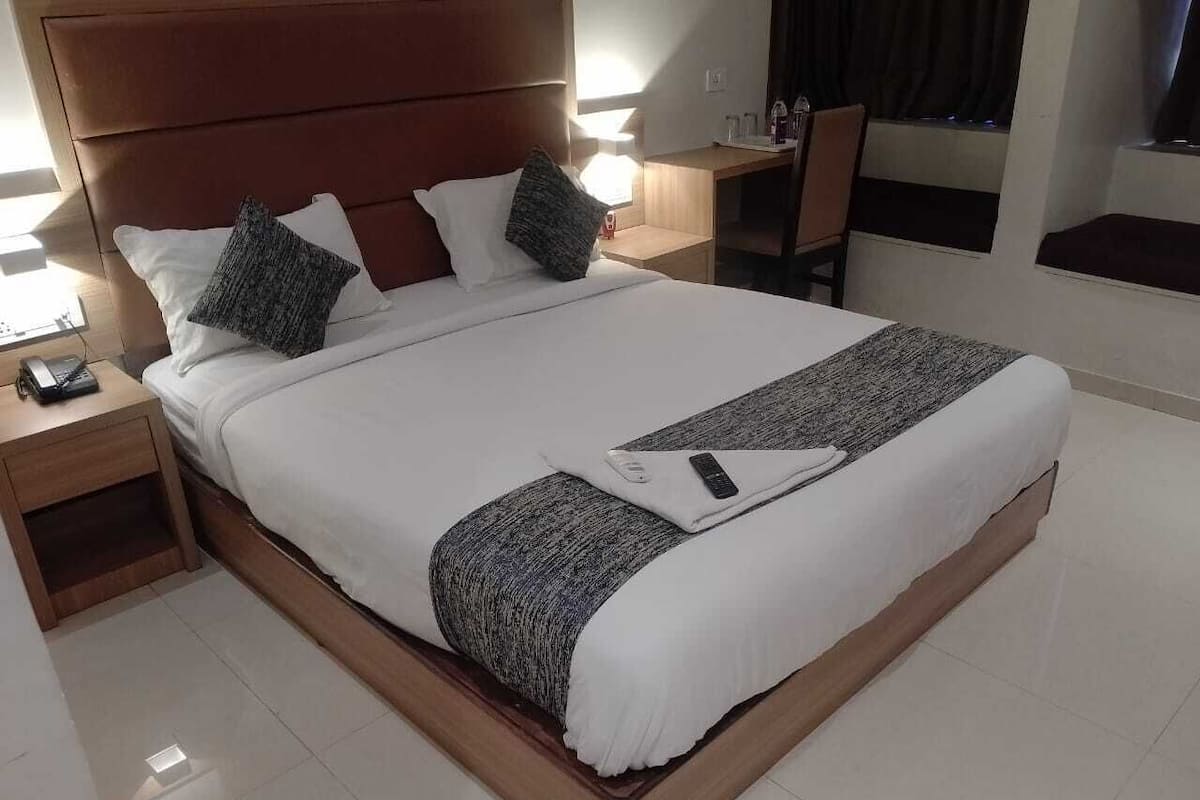 Uma cama bem arrumada com um edredom branco e duas almofadas, uma mesa de cabeceira com abajur, e uma cadeira em um quarto de hotel moderno.