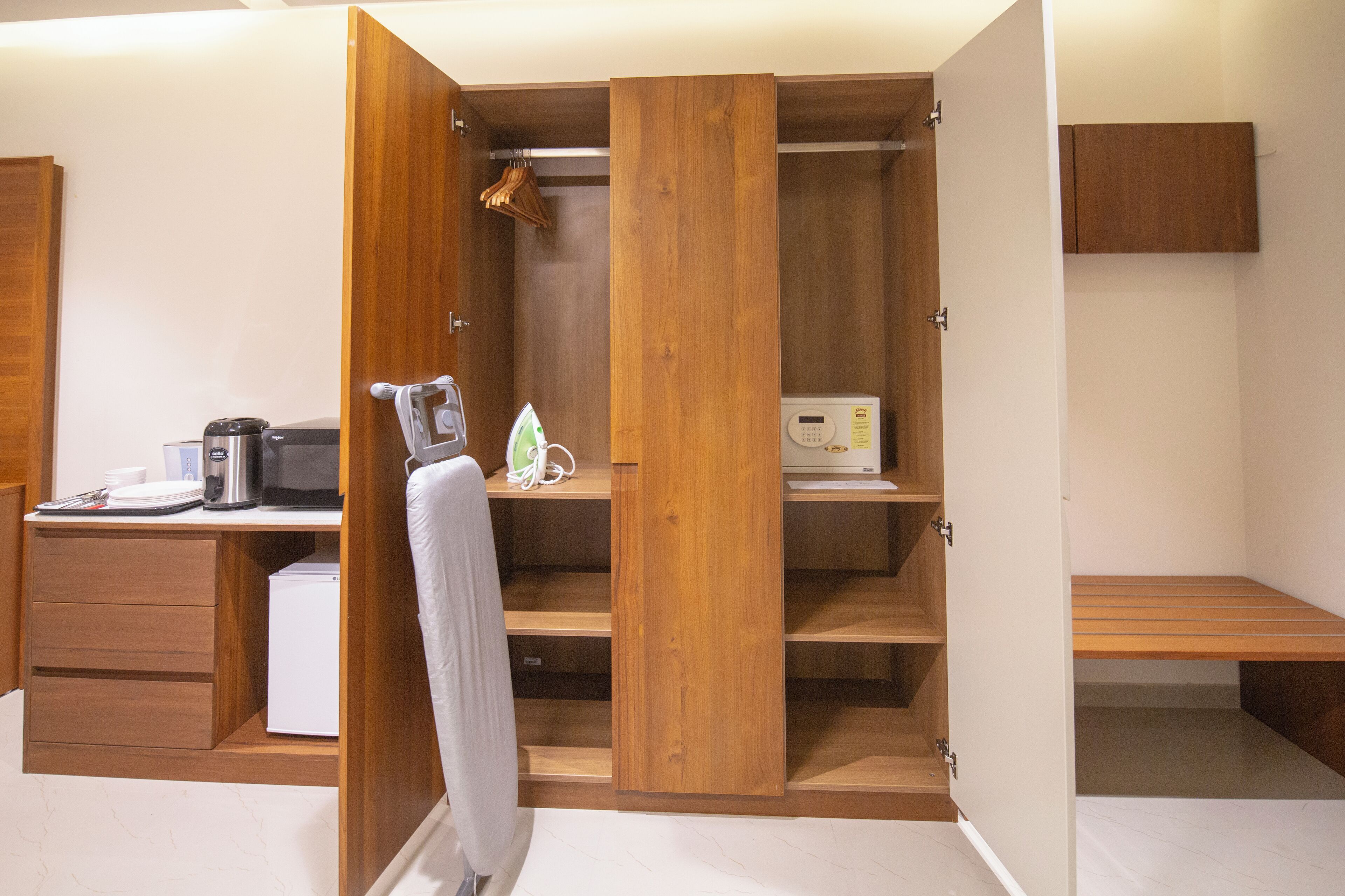 Quarto compacto com guarda-roupa de madeira, pequena mesa com chaleira, micro-ondas e cabideiro.