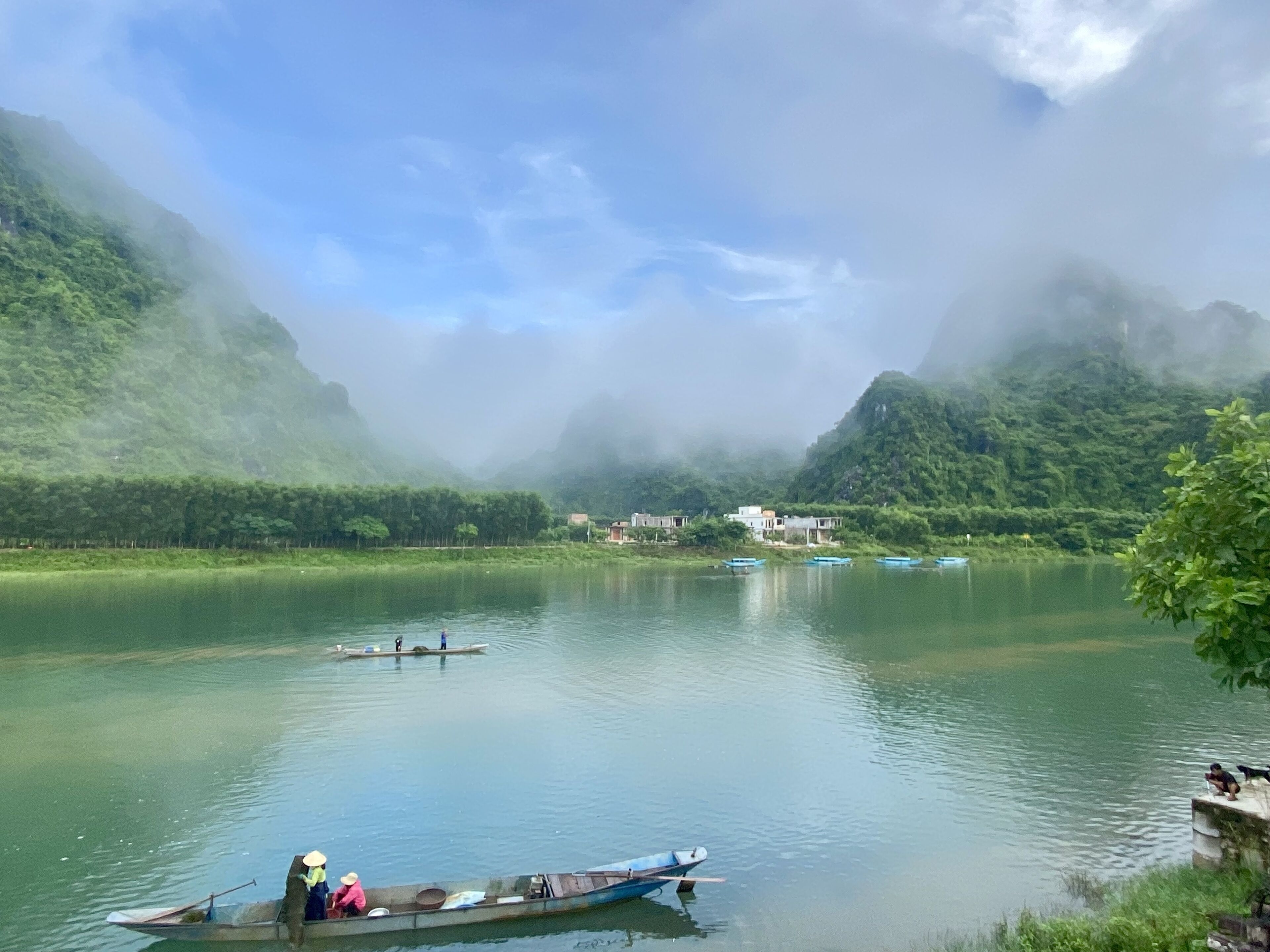 Um lago calmo com barcos, rodeado por colinas verdes exuberantes e montanhas nevoentas.
