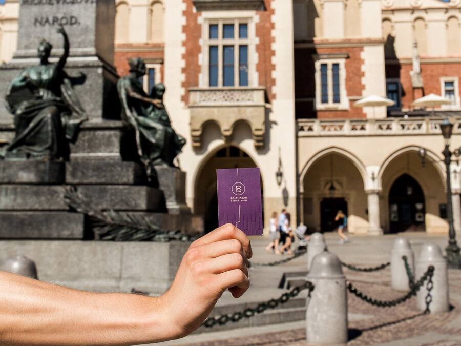 Uma mão segura um cartão roxo em frente a uma estátua e um edifício com janelas em arco e cúpula.