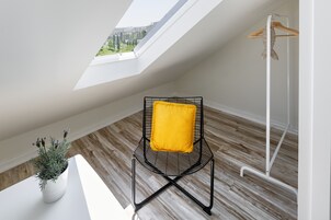 Um quarto com claraboia, uma cadeira de arame preta com almofada amarela, um cabide de madeira e um vaso com planta.