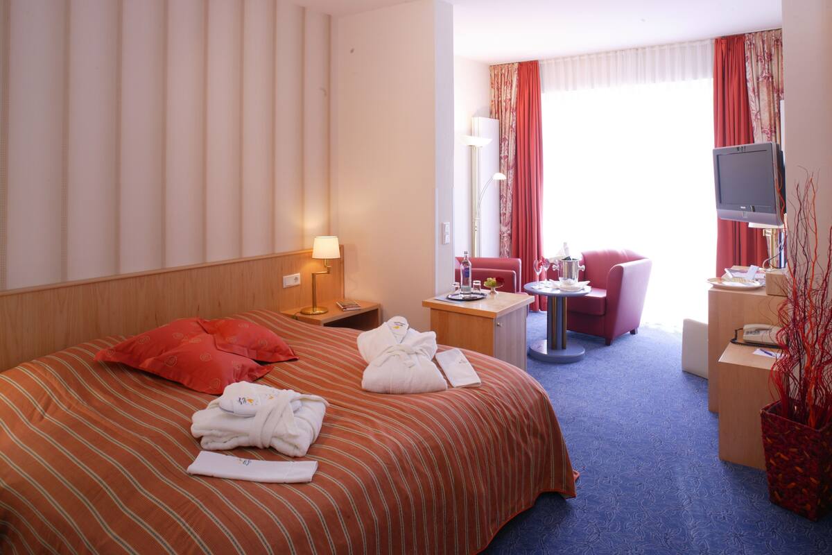 Quarto de hotel com uma cama, uma escrivaninha, uma cadeira, uma televisão e uma janela com cortinas.