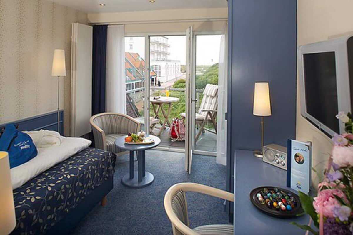 Quarto de hotel com uma cama, uma pequena mesa com um prato de comida, uma cadeira, uma varanda com mesa e cadeiras e uma televisão.