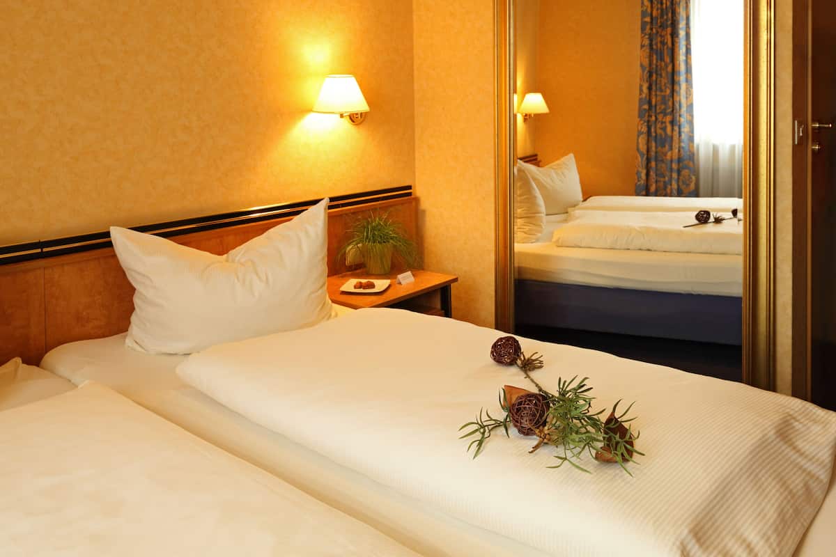 Quarto de hotel com duas camas, uma mesa de cabeceira com abajur e um espelho refletindo o quarto.