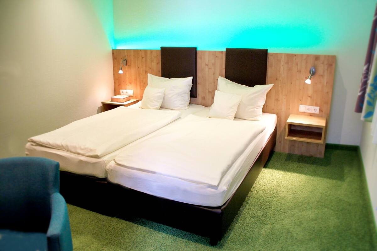 Uma cama de casal com cabeceira de madeira, duas mesas de cabeceira com abajures e um tapete verde.