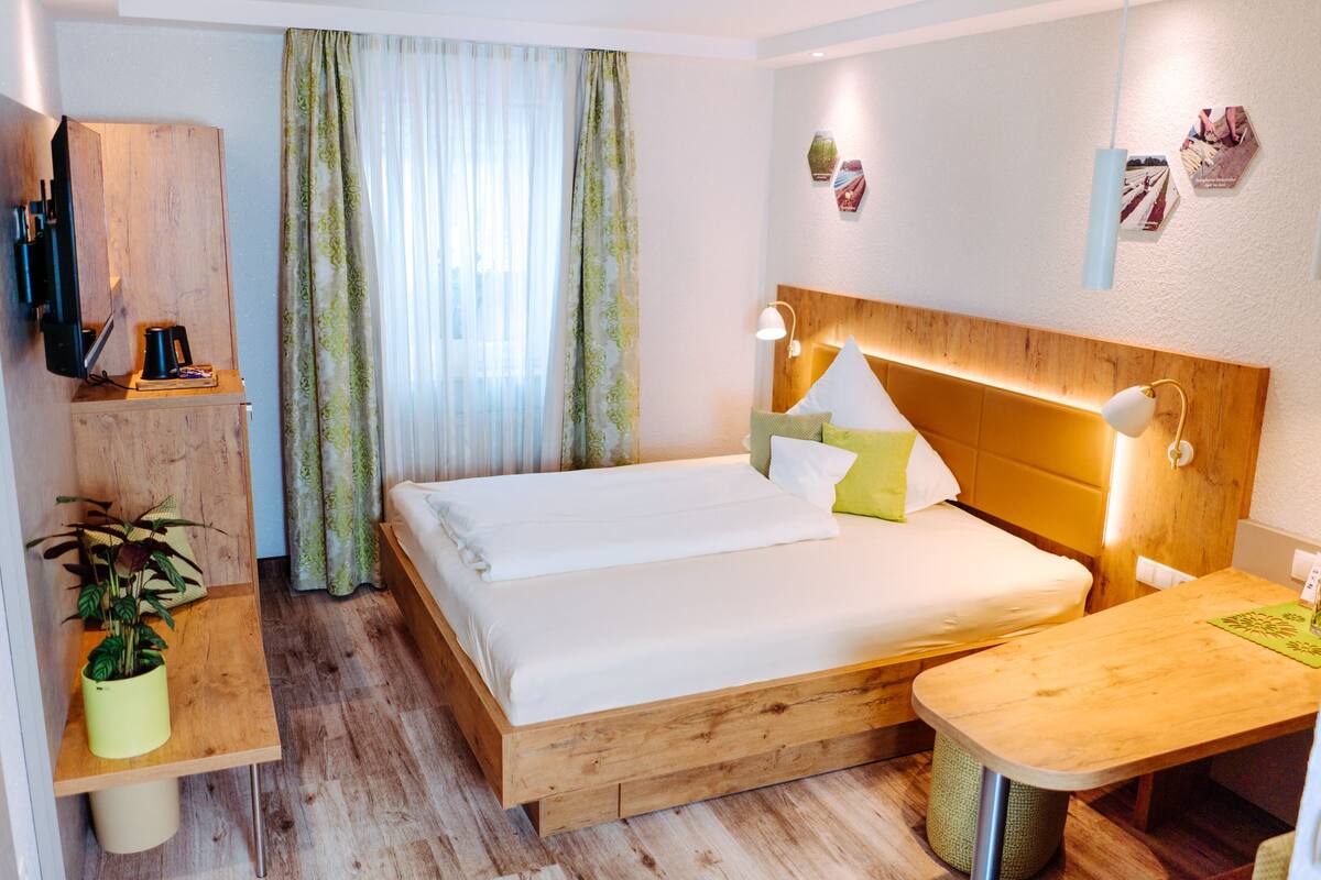 Quarto de hotel com cama de madeira, luminárias de cabeceira, uma mesinha, televisão e planta em vaso.