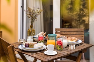 Mesa de madeira posta com itens de café da manhã, incluindo pão, frutas e bebidas, com um vaso de flores e saleiros em um pátio ensolarado.