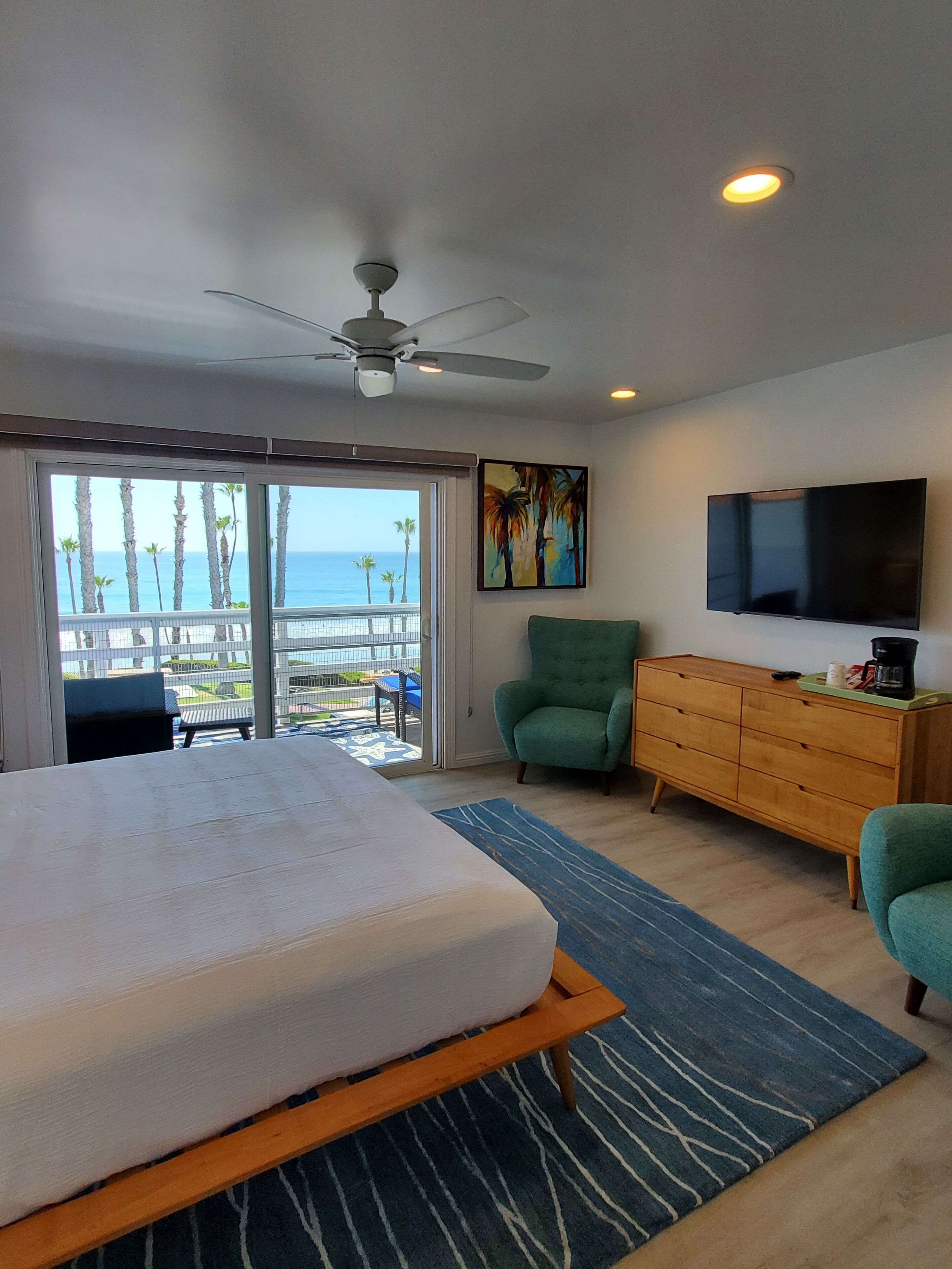 Quarto de hotel com uma cama, uma poltrona verde, penteadeira de madeira, televisão de tela plana e varanda com vista para palmeiras e o oceano.