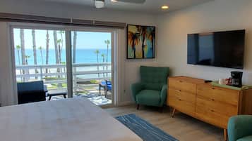 Quarto de hotel com uma cama, uma poltrona verde, penteadeira de madeira, televisão de tela plana e varanda com vista para palmeiras e o oceano.