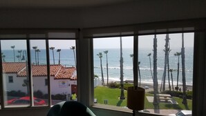 Quarto com uma grande janela com vista para uma praia com palmeiras e um edifício com telhado de telhas vermelhas.