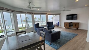 Ampla sala de estar com mesa de jantar, cadeiras, sofá e televisão. O quarto possui janelas grandes com vista para o oceano e palmeiras.
