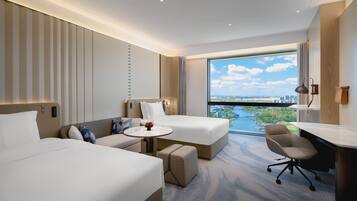 Quarto de hotel com duas camas, uma mesa pequena, uma escrivaninha e vista para a cidade e para o rio.