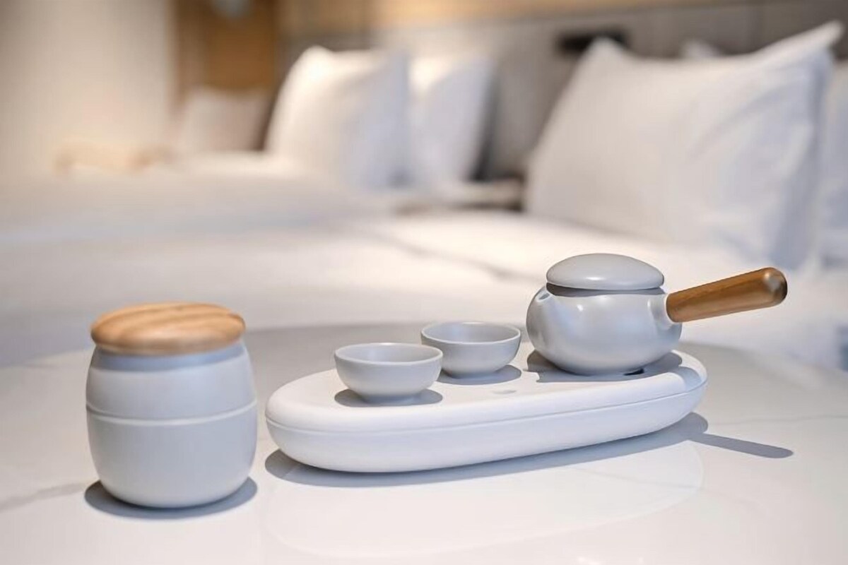 Um conjunto de chá bem organizado sobre uma mesa branca num quarto de hotel, com uma cama ao fundo.