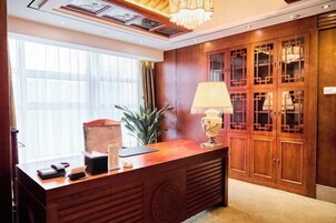 Balcão de recepção com telefone, luminária e planta. Há uma janela grande com cortinas, e armários de madeira com portas de vidro.