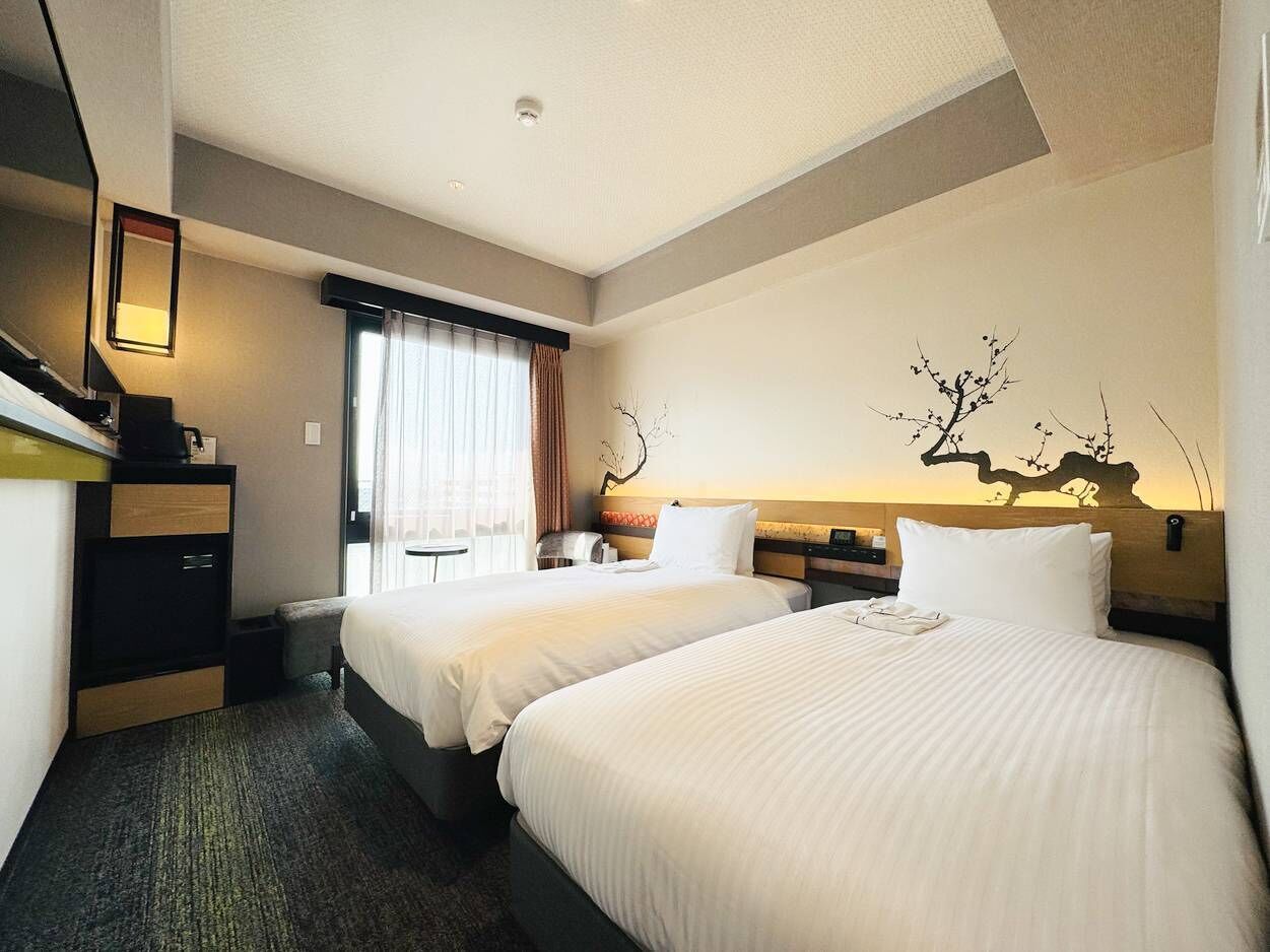 Quarto de hotel com duas camas, uma escrivaninha, uma cadeira e uma janela com cortinas.
