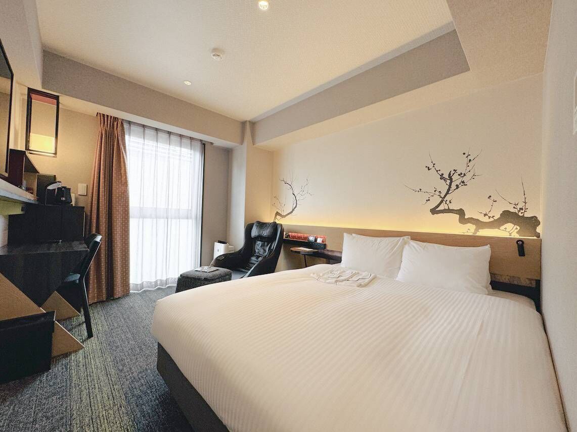 Quarto de hotel com cama, escrivaninha, cadeira, mesa de cabeceira e uma obra de arte na parede.