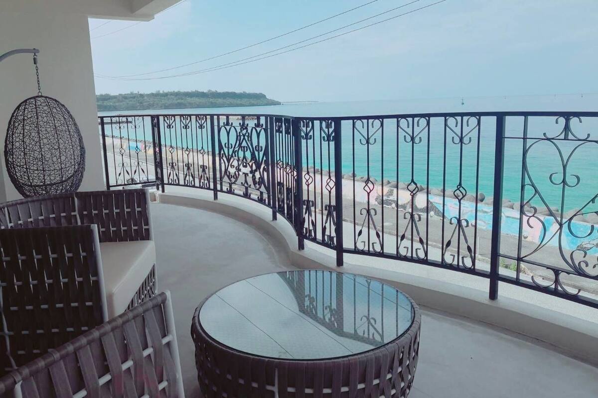 Varanda com mesa e cadeiras de vidro, com vista para uma praia e uma visão clara do mar e da terra ao longe.