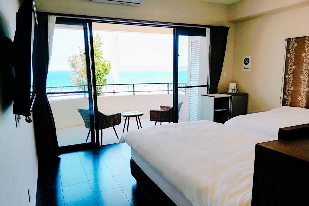 Quarto de hotel com uma cama, uma mesa pequena, uma cadeira e vista para o oceano por uma porta de correr.