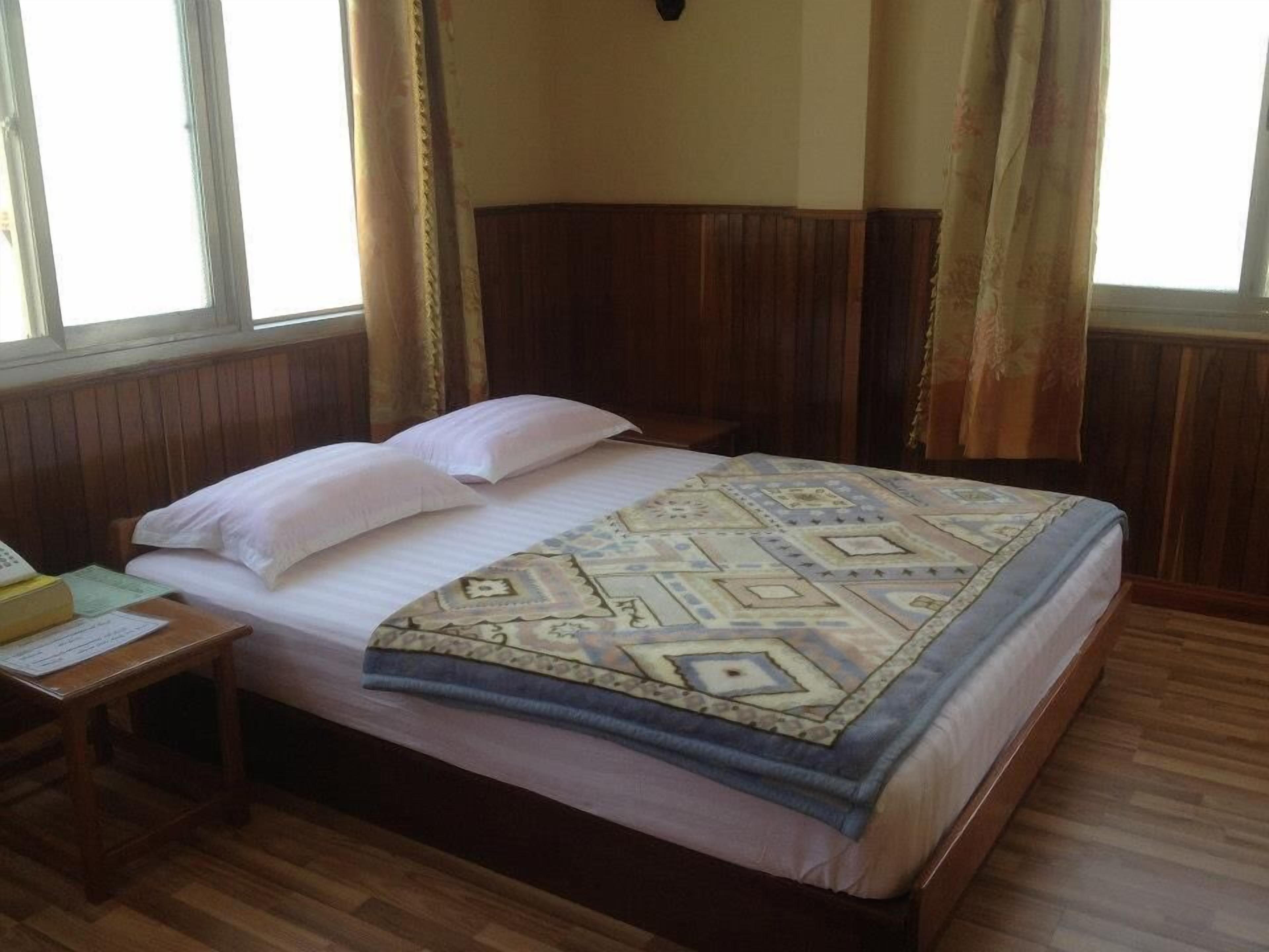 Quarto com cabeceira de madeira, cama com roupa de cama branca e manta estampada, mesa de cabeceira com abajur e janela com cortinas.