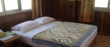 Quarto com cabeceira de madeira, cama com roupa de cama branca e manta estampada, mesa de cabeceira com abajur e janela com cortinas.