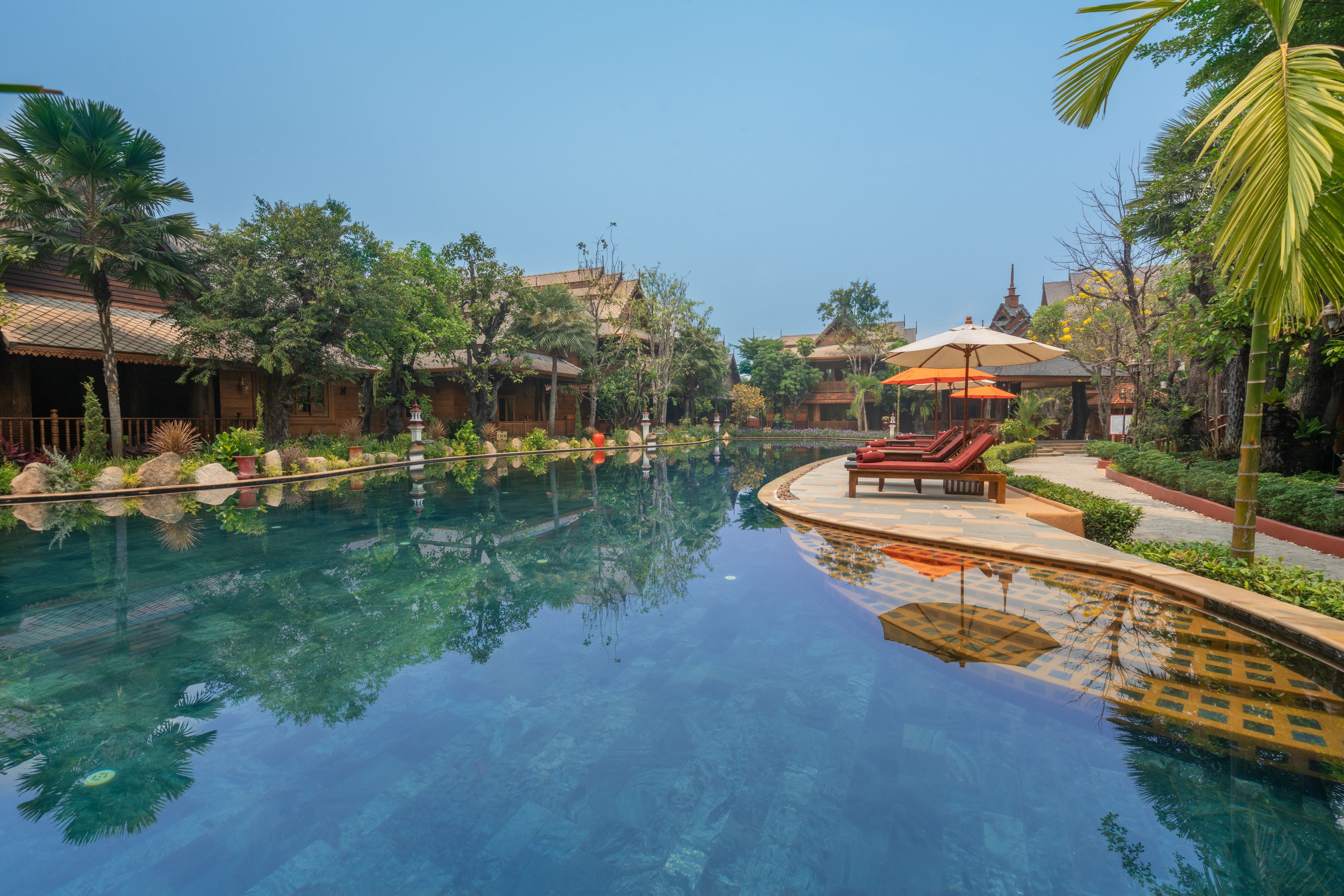 Piscina límpida com espreguiçadeiras e guarda-sóis, rodeada por vegetação tropical e arquitetura tradicional.