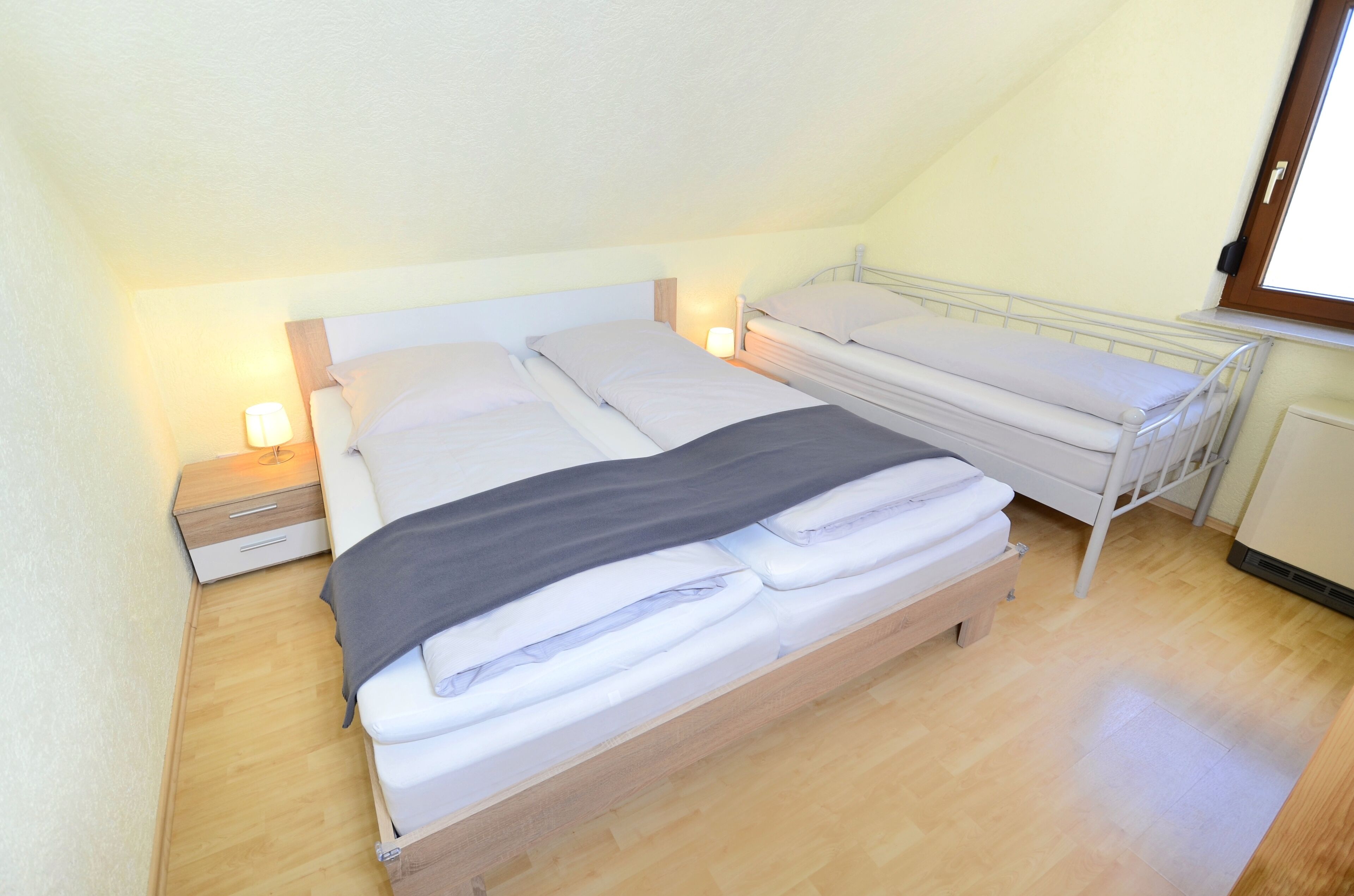 Uma cama de solteiro com cabeceira de madeira, uma mesa de cabeceira com abajur e uma janela com vista para o exterior.