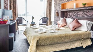 Quarto de hotel com cama, duas cadeiras, escrivaninha com computador e janela com cortinas.
