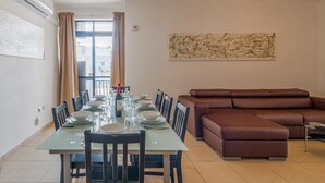 Uma área de refeições com uma mesa comprida posta para seis pessoas, rodeada por cadeiras, um sofá marrom e um grande quadro abstrato na parede.