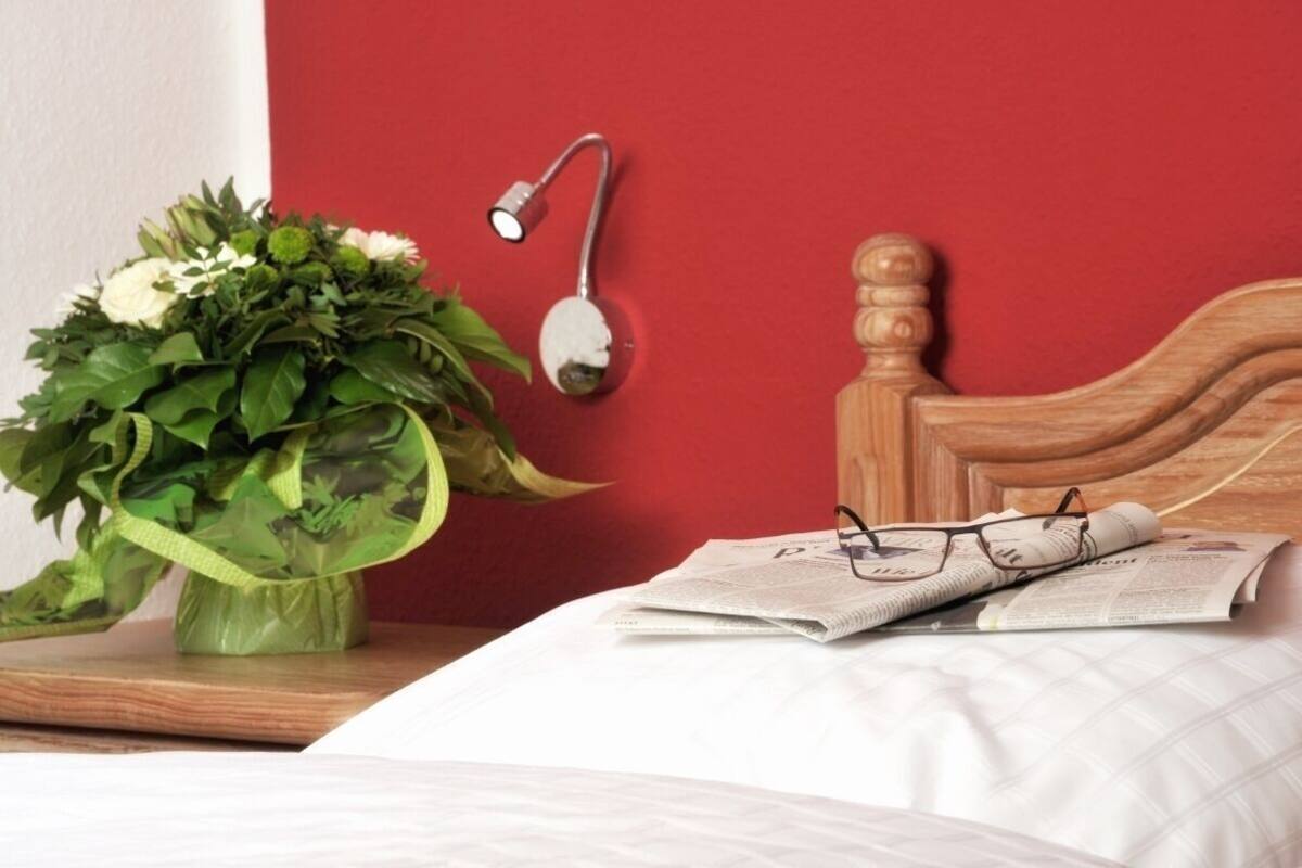 Cama bem arrumada com jornal, óculos e um vaso de flores sobre uma mesa de cabeceira, em frente a uma parede vermelha.