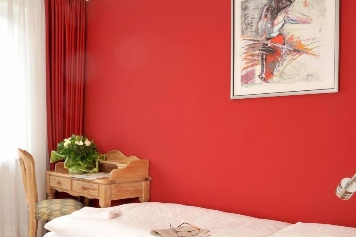 Quarto com parede vermelha, cama de madeira, escrivaninha com cadeira e um quadro.