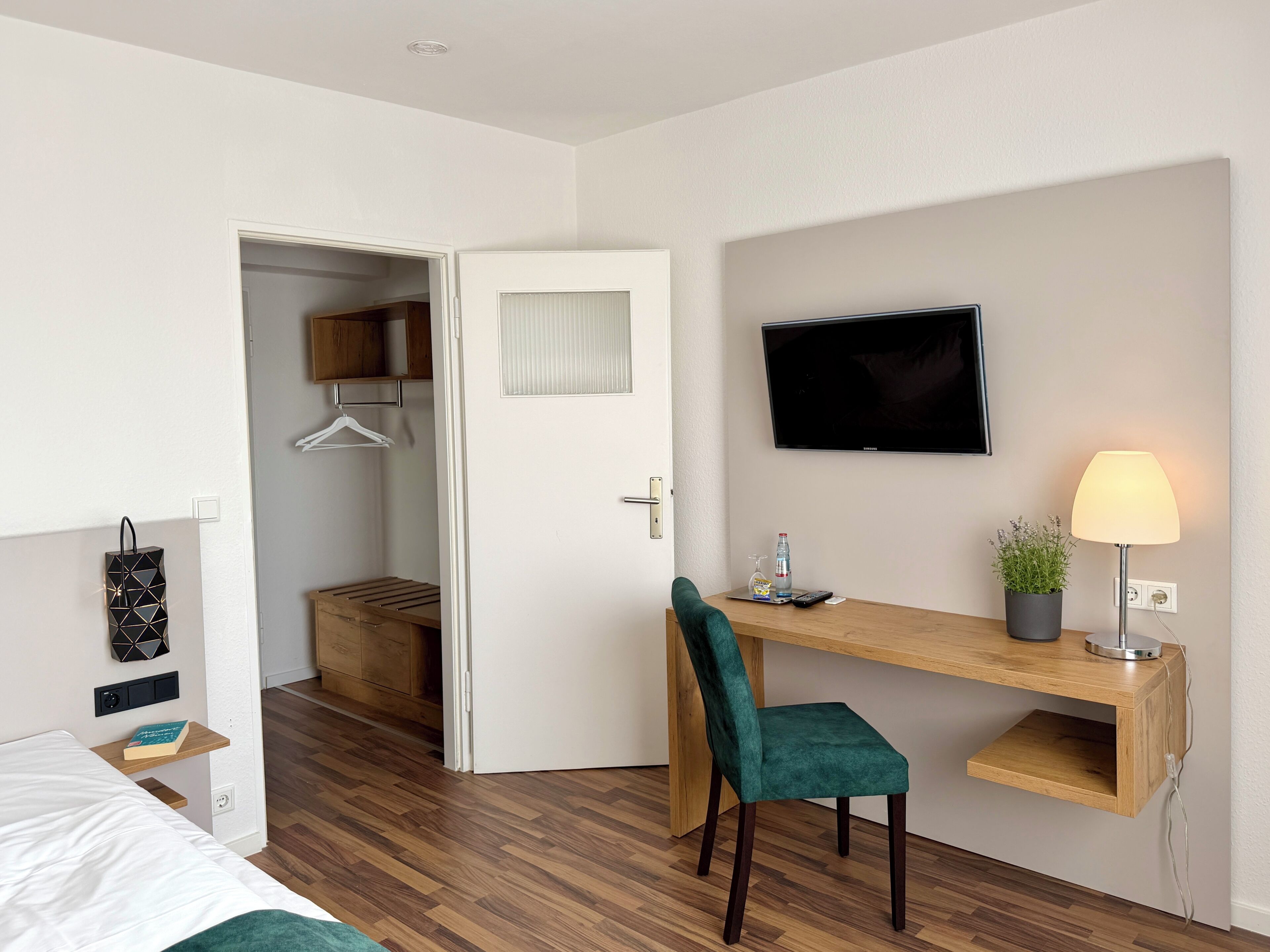 Quarto de hotel com cama, escrivaninha com cadeira, televisão, luminária, planta e armário.
