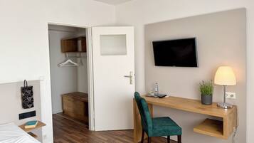 Quarto de hotel com cama, escrivaninha com cadeira, televisão, luminária, planta e armário.