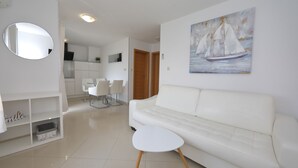 Uma sala de estar moderna com um sofá branco, uma mesinha pequena branca, um espelho redondo e uma pintura de veleiros.