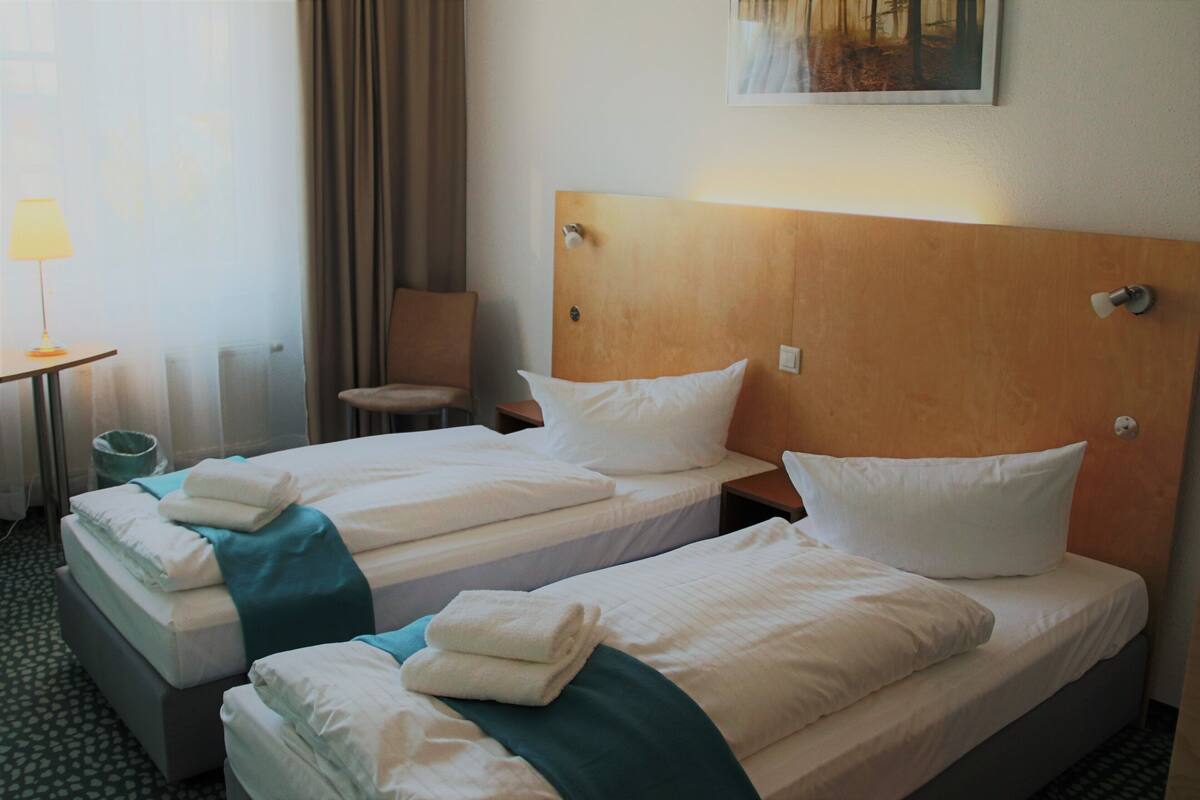 Quarto de hotel com duas camas de solteiro, uma mesinha com luminária, uma cadeira e um quadro na parede.