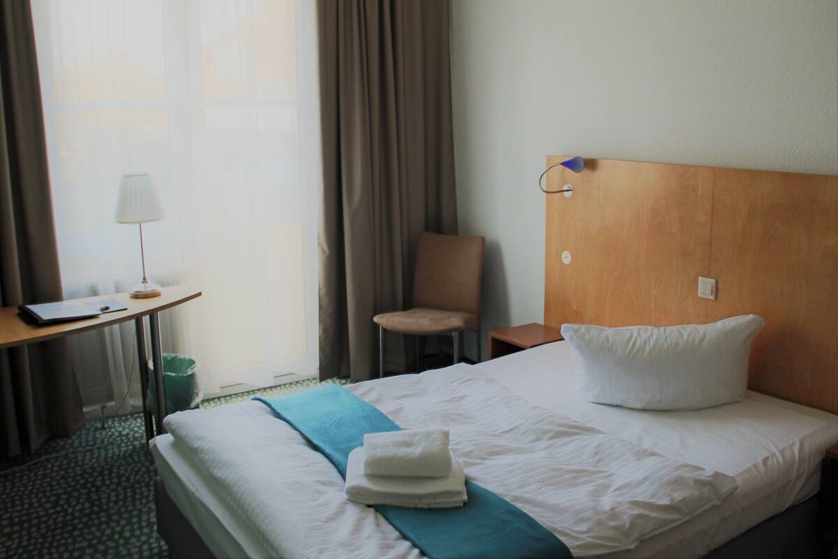 Quarto de hotel com cama, escrivaninha, cadeira, luminária e janela com cortinas.