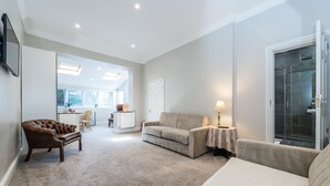 Uma sala de estar ampla com um sofá de couro marrom, um sofá bege e um sofá branco. Há uma mesinha com uma luminária, um radiador e uma porta que dá para um espaço com chuveiro.