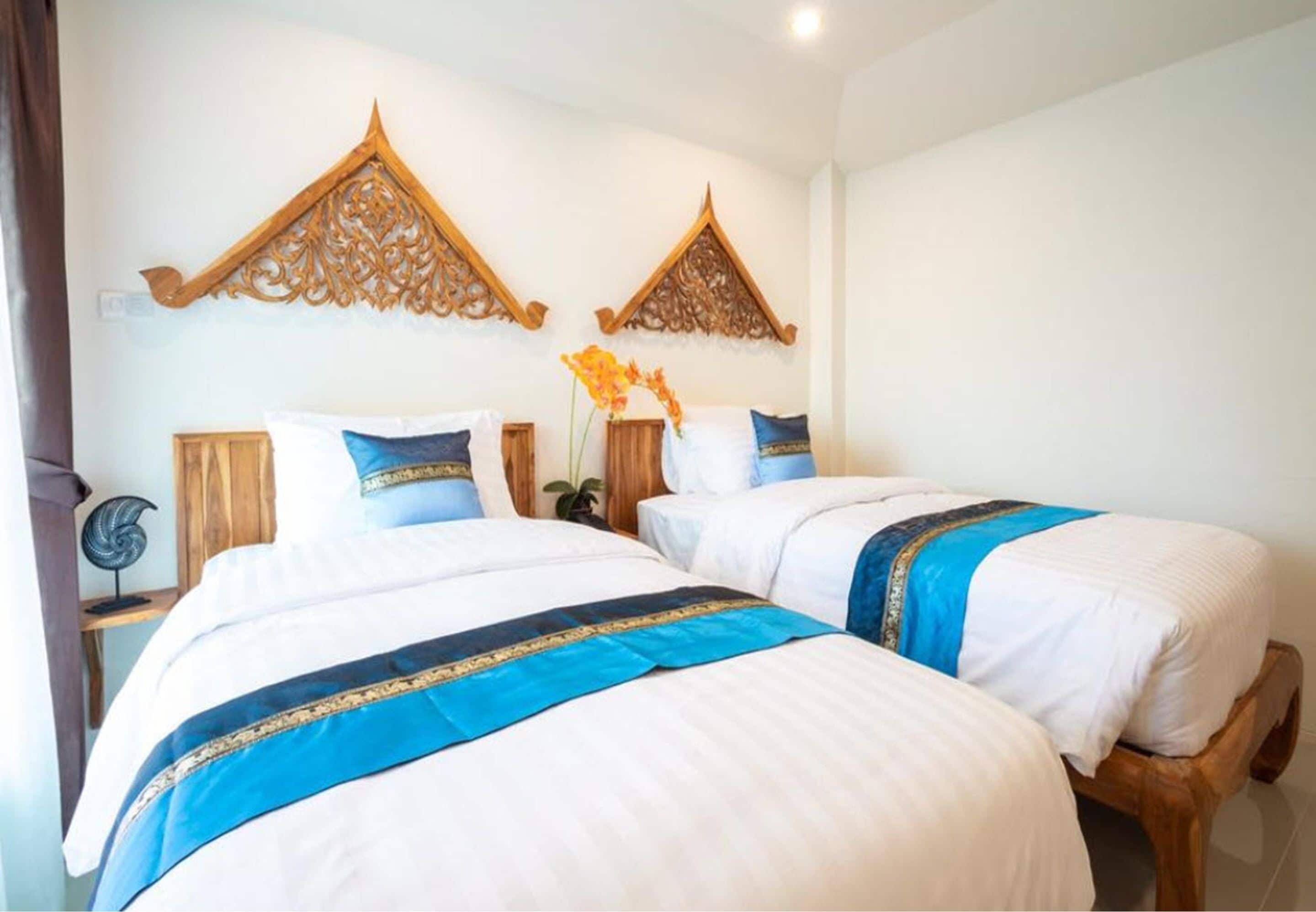Quarto de hotel com duas camas, cada uma com roupa de cama branca e detalhes azuis. Há cabeceiras de madeira e um elemento decorativo acima das camas.