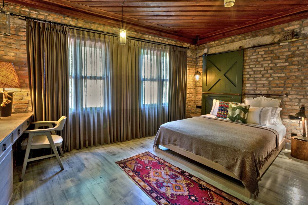 Quarto com cama, escrivaninha, cadeira e uma janela grande com cortinas.