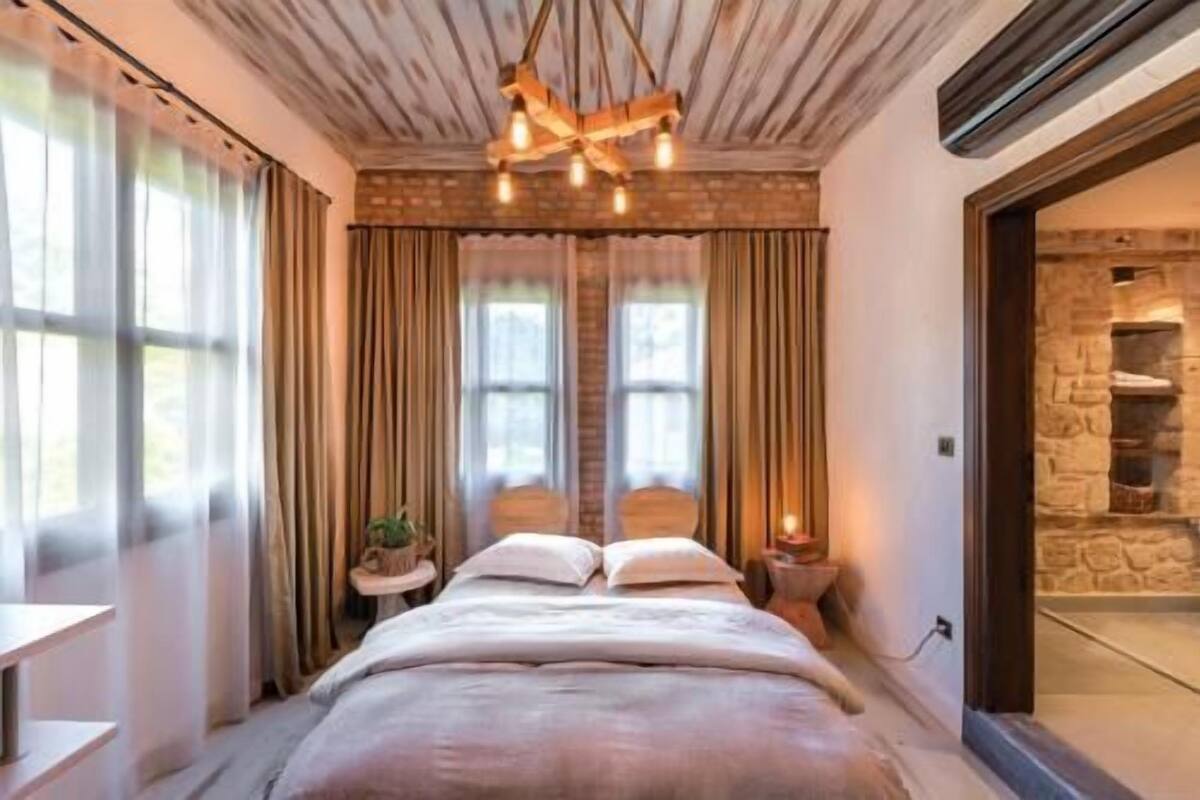 Quarto com cama, cortinas, luminária pendente e parede de pedra.